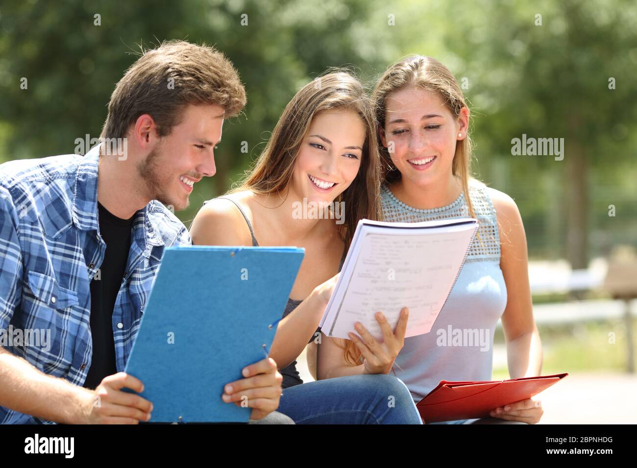 Three happy students studying together comparing notes in a campus park ...