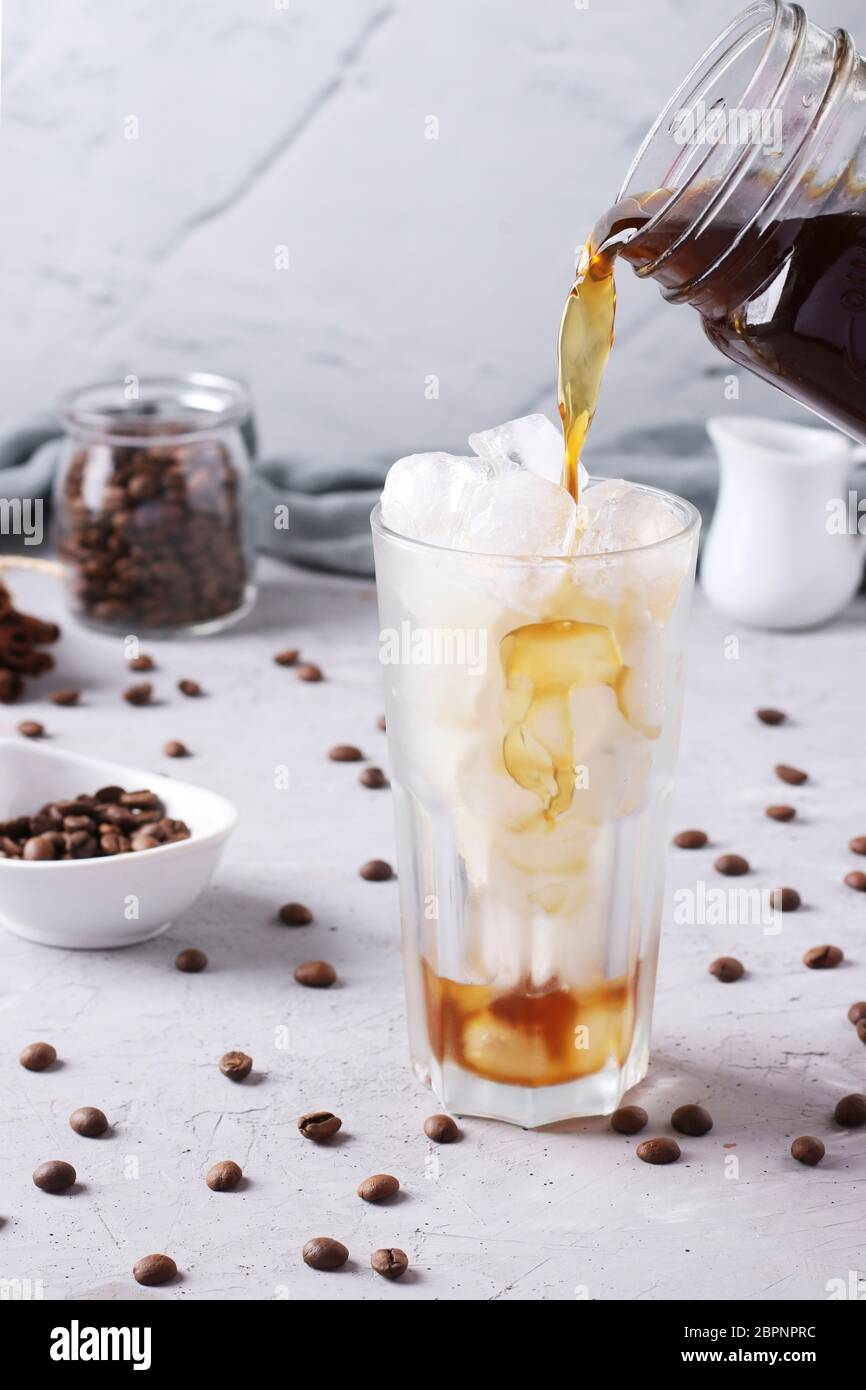 Cold coffee is poured into a tall glass with ice cubes on light gray ...