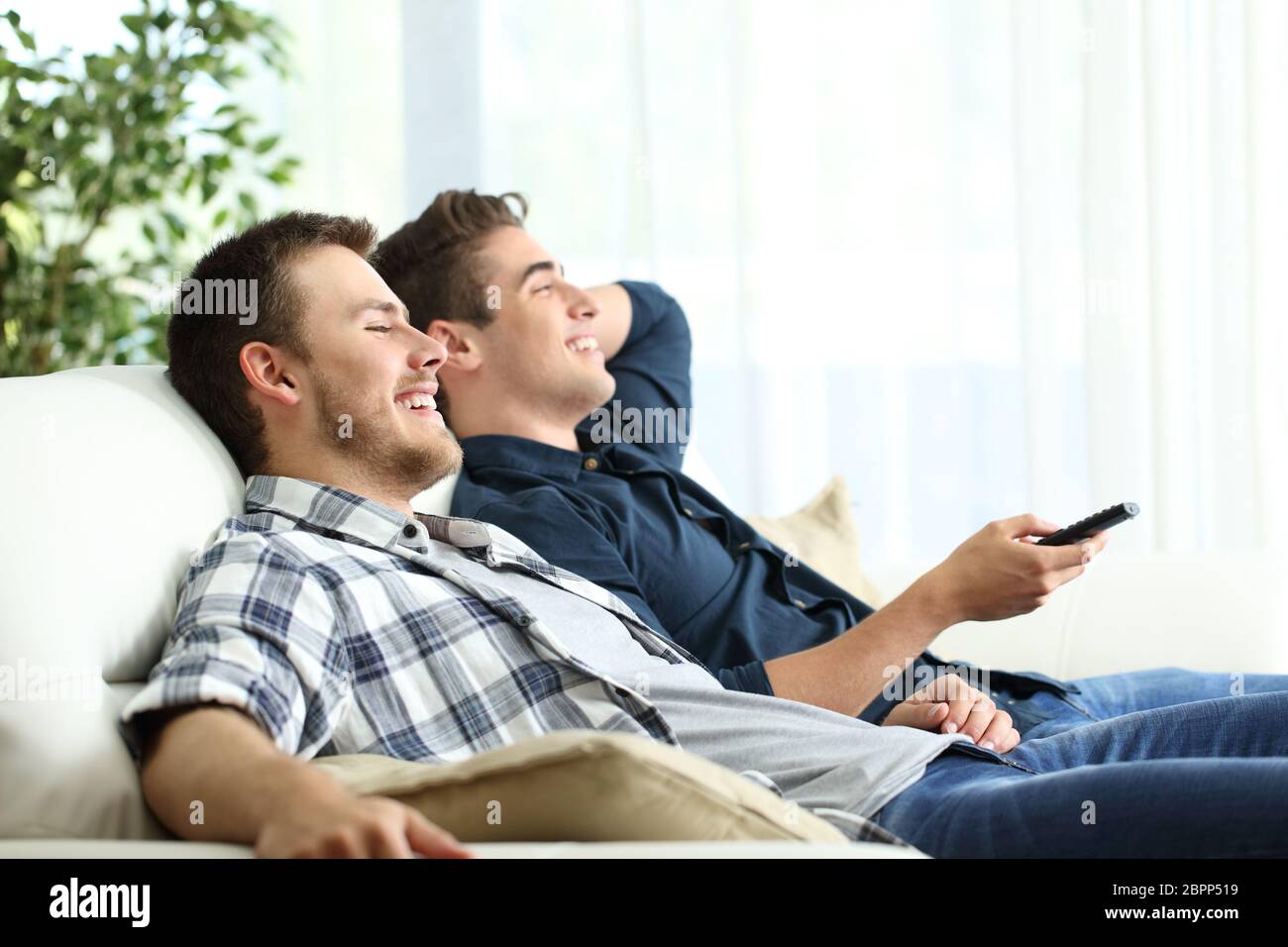 Two happy friends watching tv relaxing sitting on a couch in the living ...