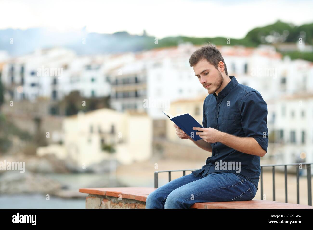 Serious man reading a paper book sitting on a ledge in a coast town ...