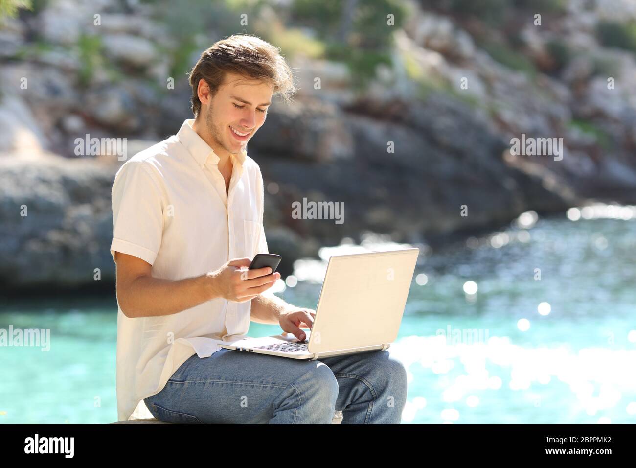 Happy man using a smart phone and a laptop on vacation on the beach ...