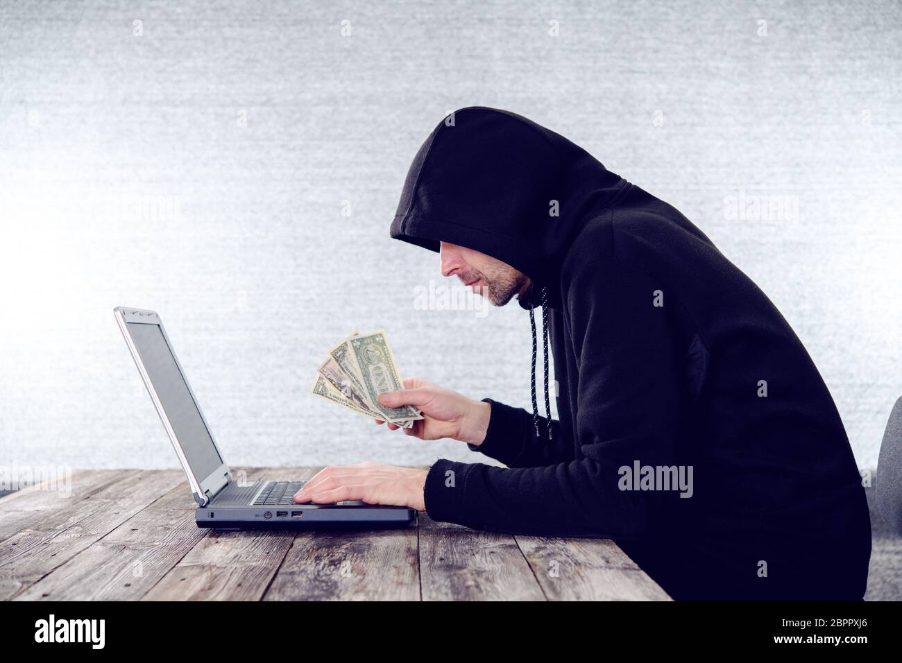 hacker in black hoody with laptop and dollar notes in front of gray ...