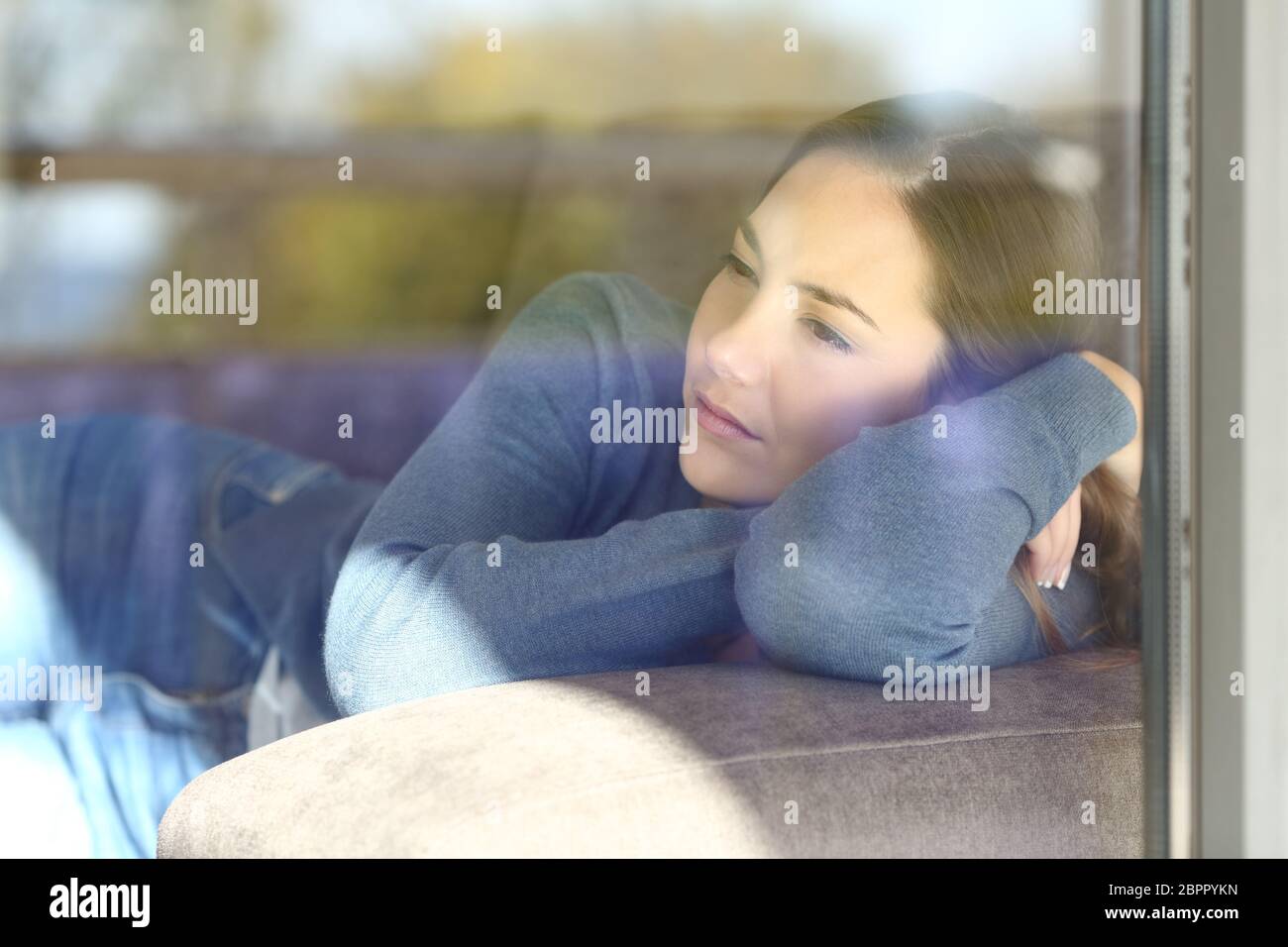 Sad woman looking through a window lying on a couch in a house interior ...
