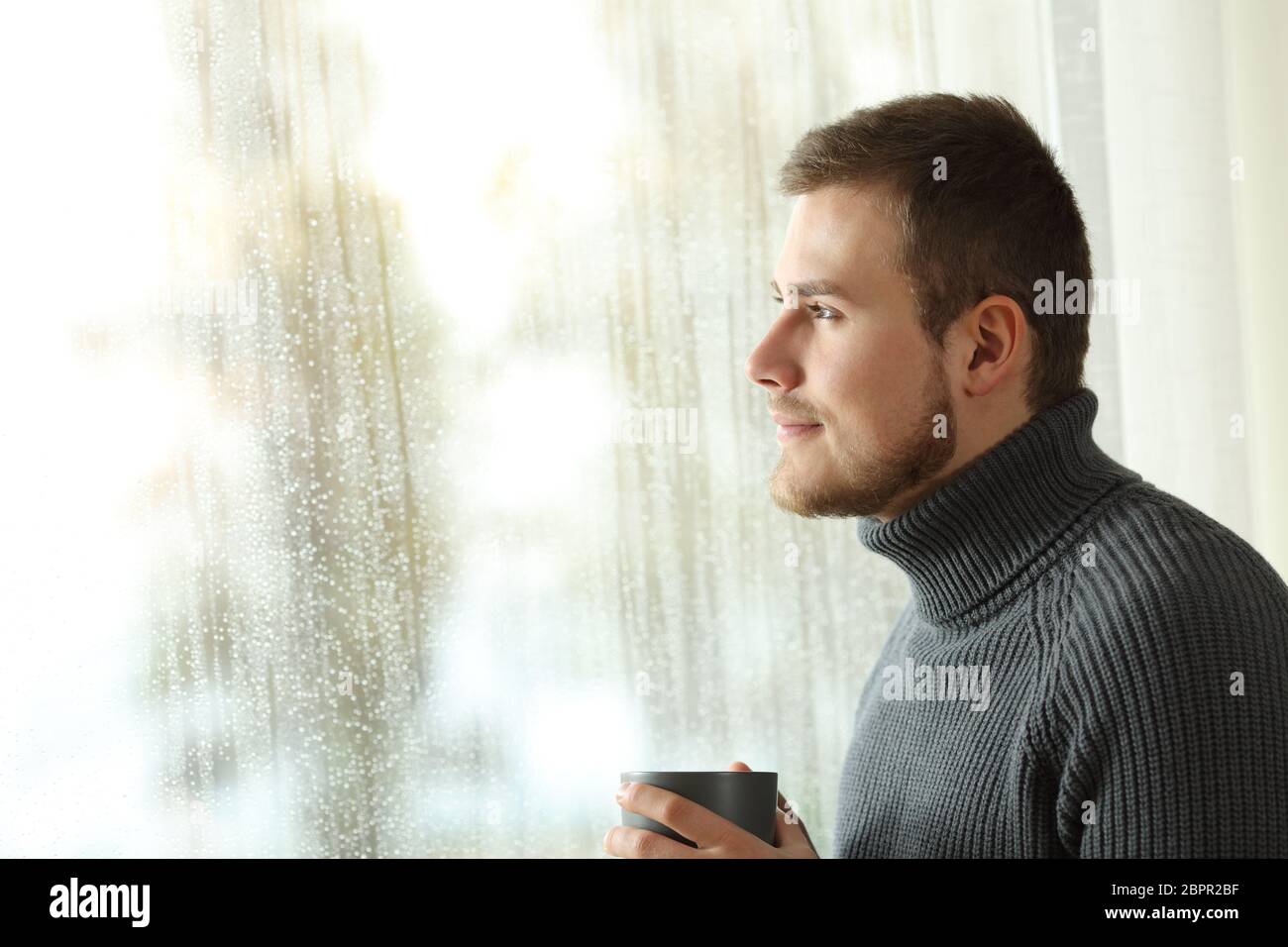 Happy man looking outside through a window at home in a rainy day Stock ...