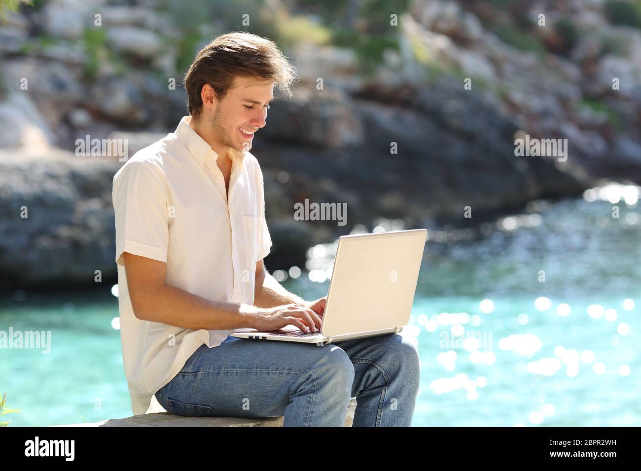 Happy guy uses a laptop writing email on summer vacation on the beach ...