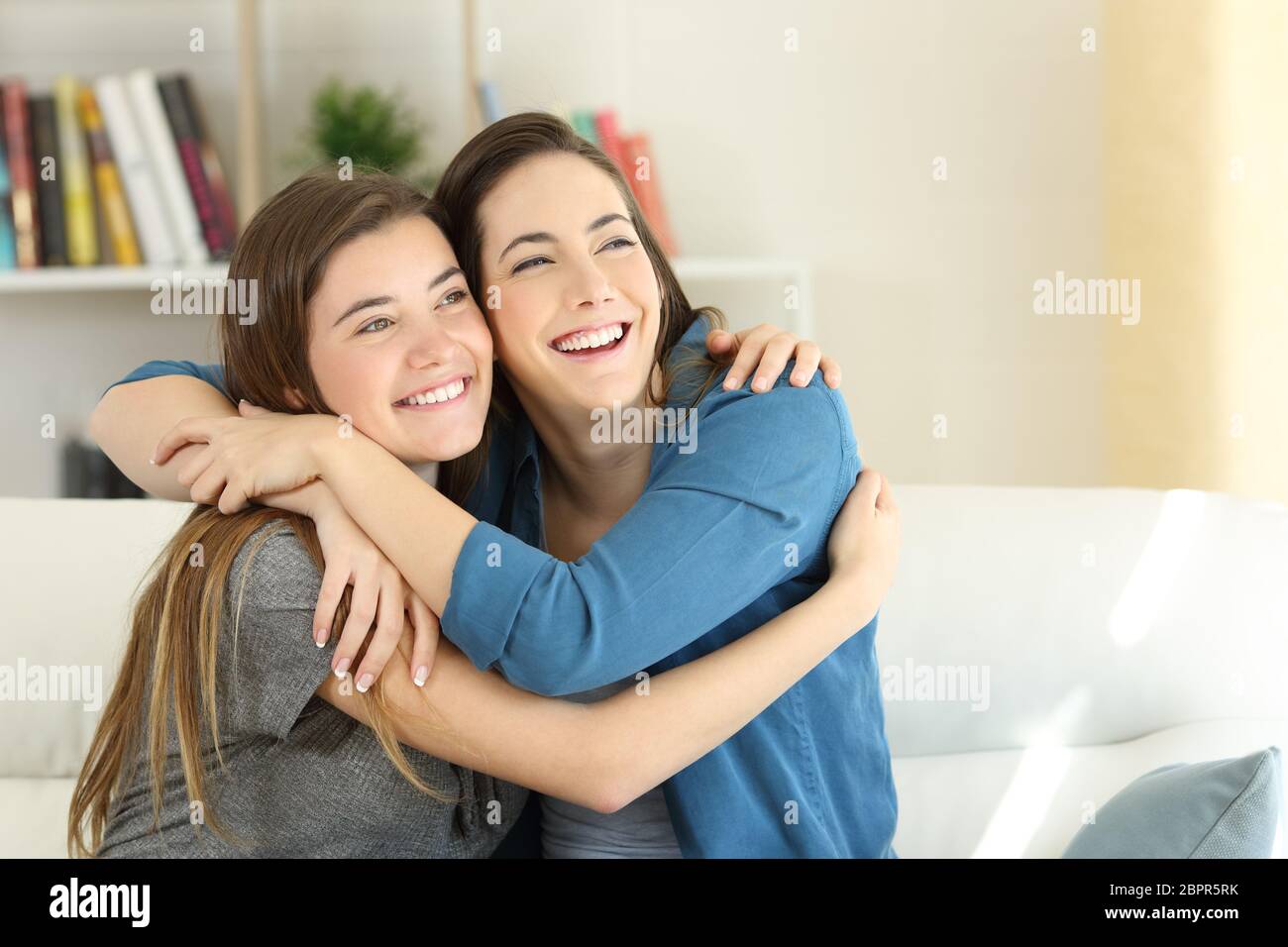 Two happy friends or sisters hugging sitting on a couch in the living ...