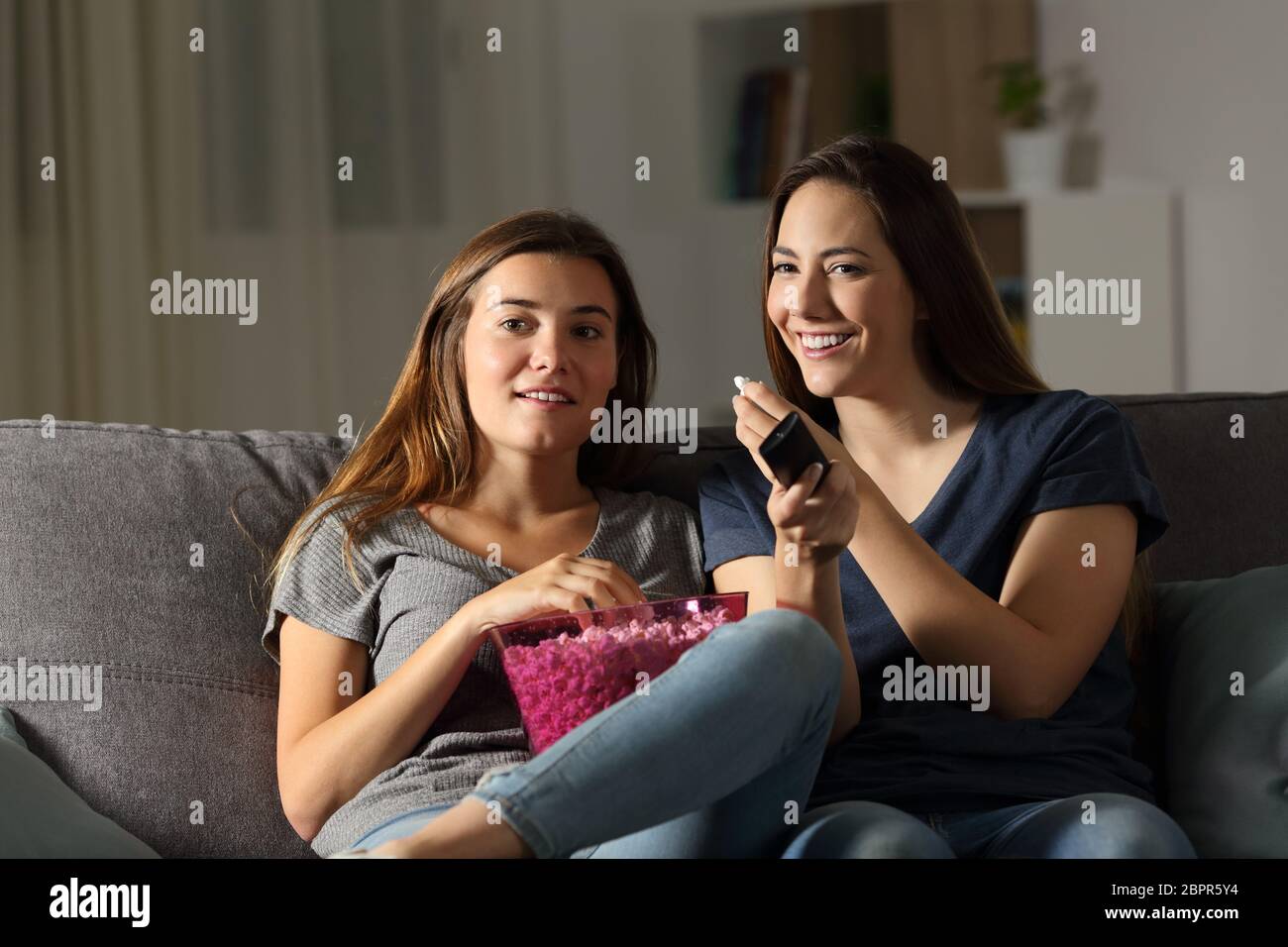 Two happy roommates wathing tv in the night sitting on a couch in the ...