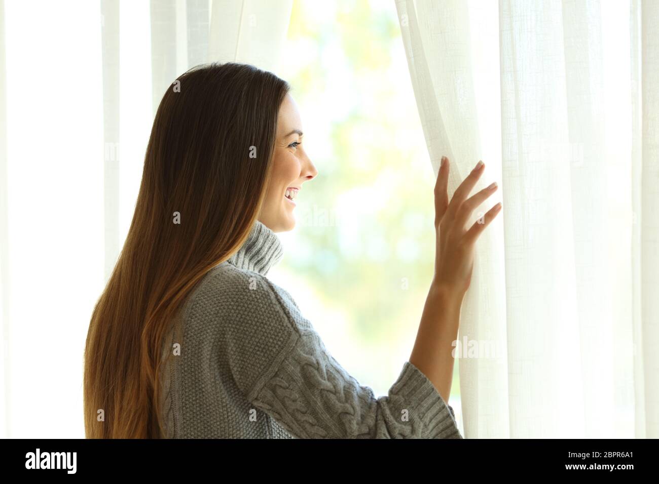 Side view of a happy woman opening curtains of a window and enjoying a ...