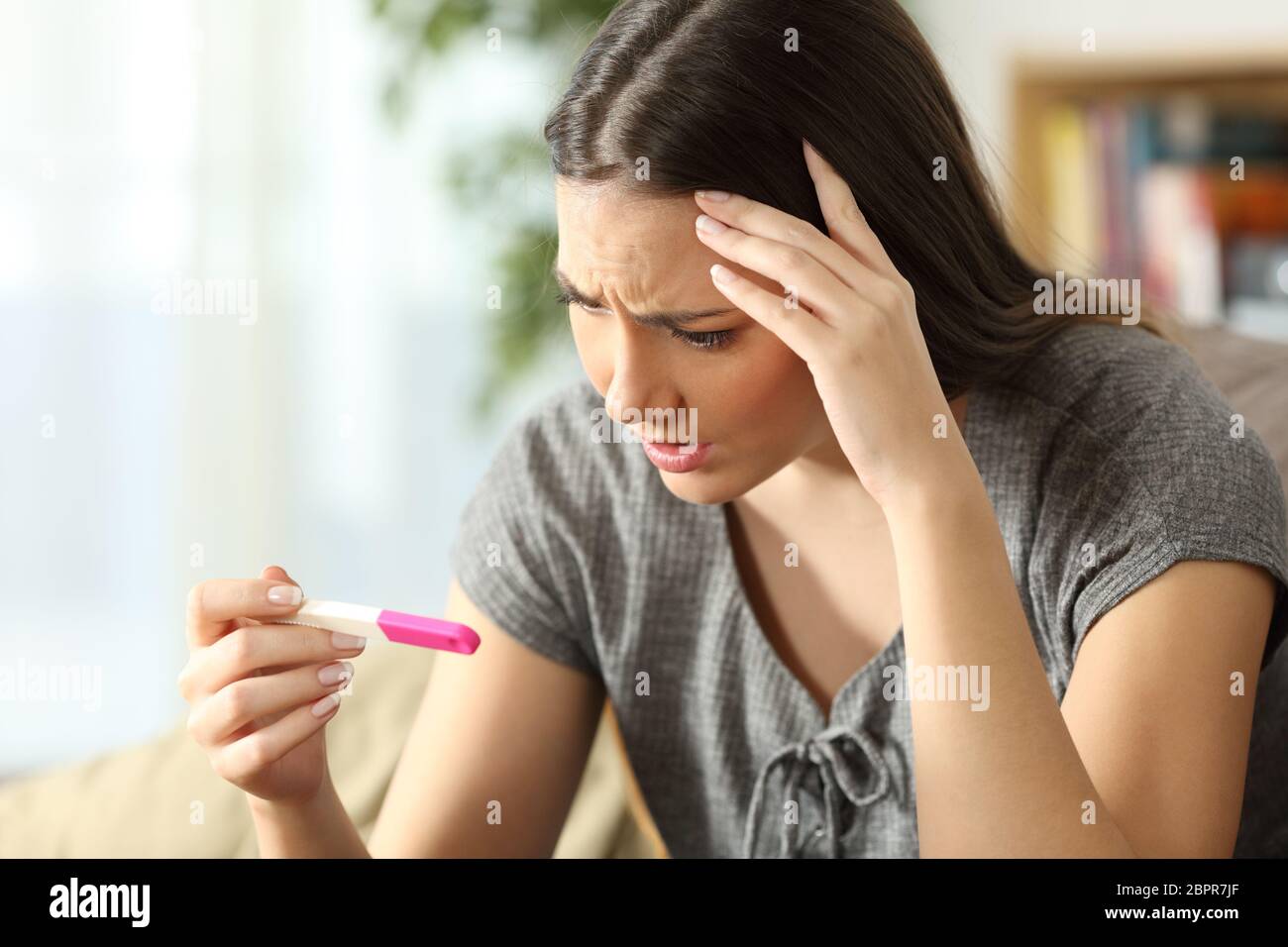 Worried woman checking a pregnancy test sitting on a couch in the ...