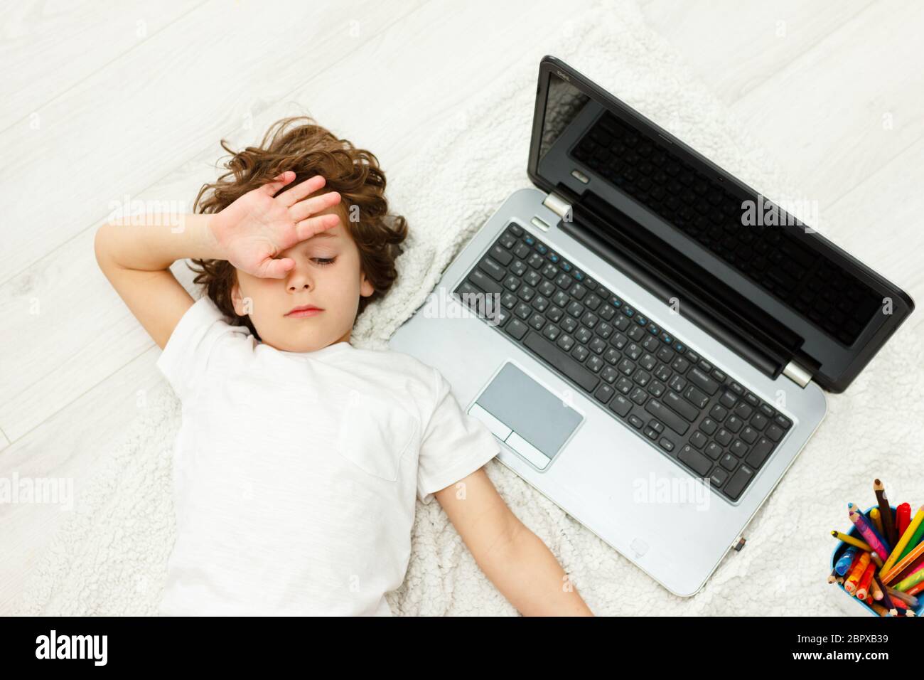 Curly boy is engaged at computer. Tired child lies covering his face ...