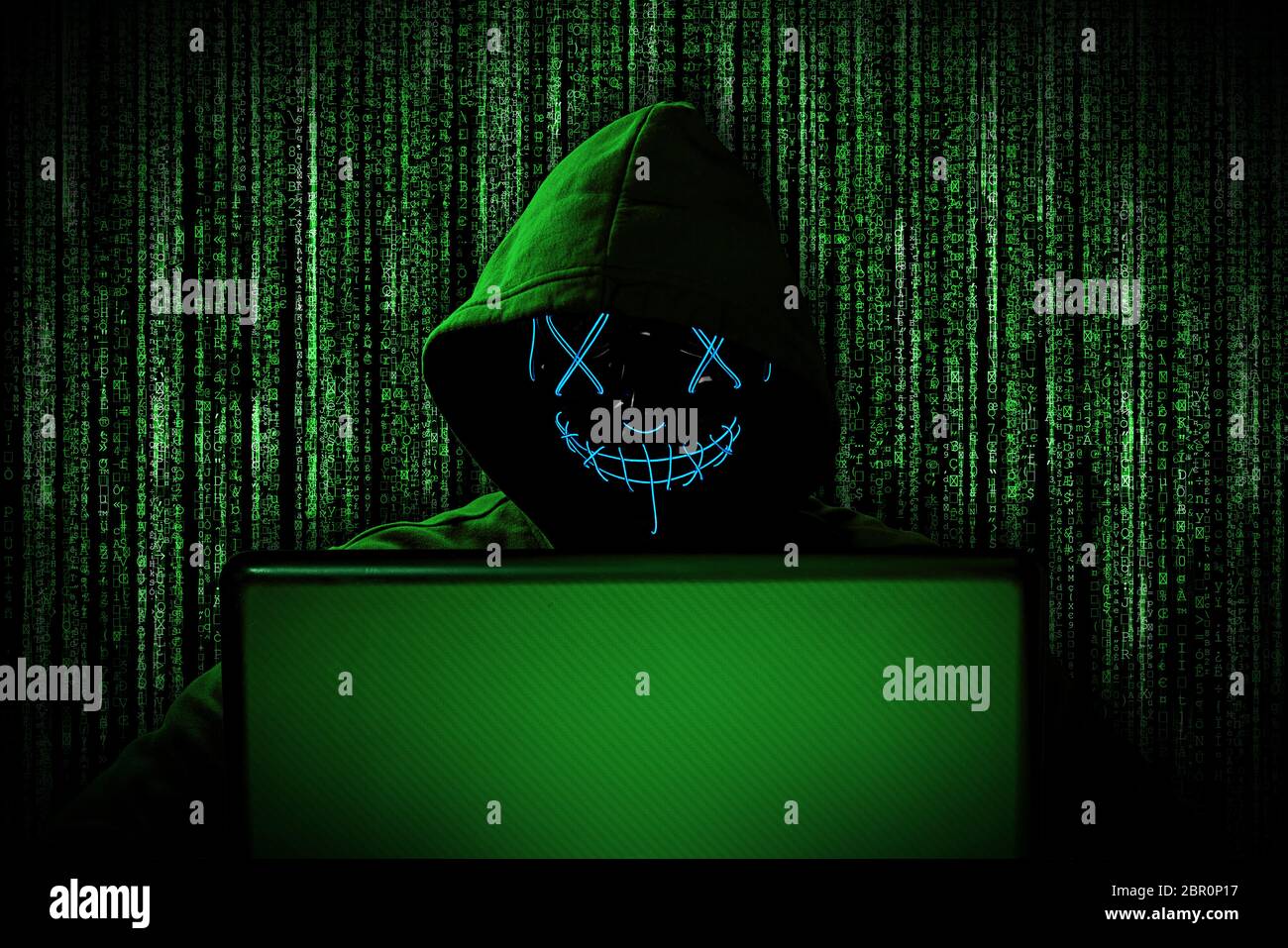 hacker with glowing mask behind notebook laptop in front of green source binary code background ...