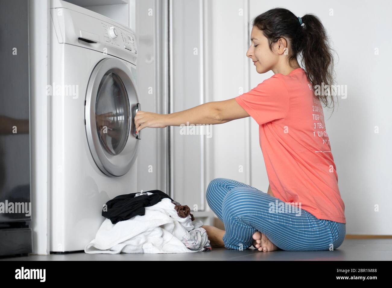 A girl loads dirty laundry into a washing machine while sitting on the ...