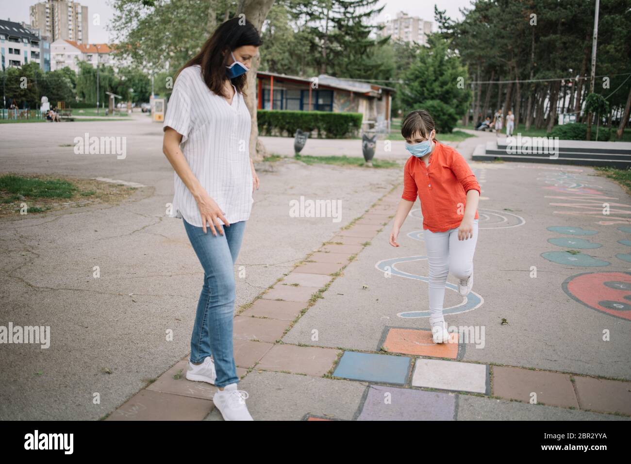 Brunette woman watching girl playing games on asphalt with masks on ...
