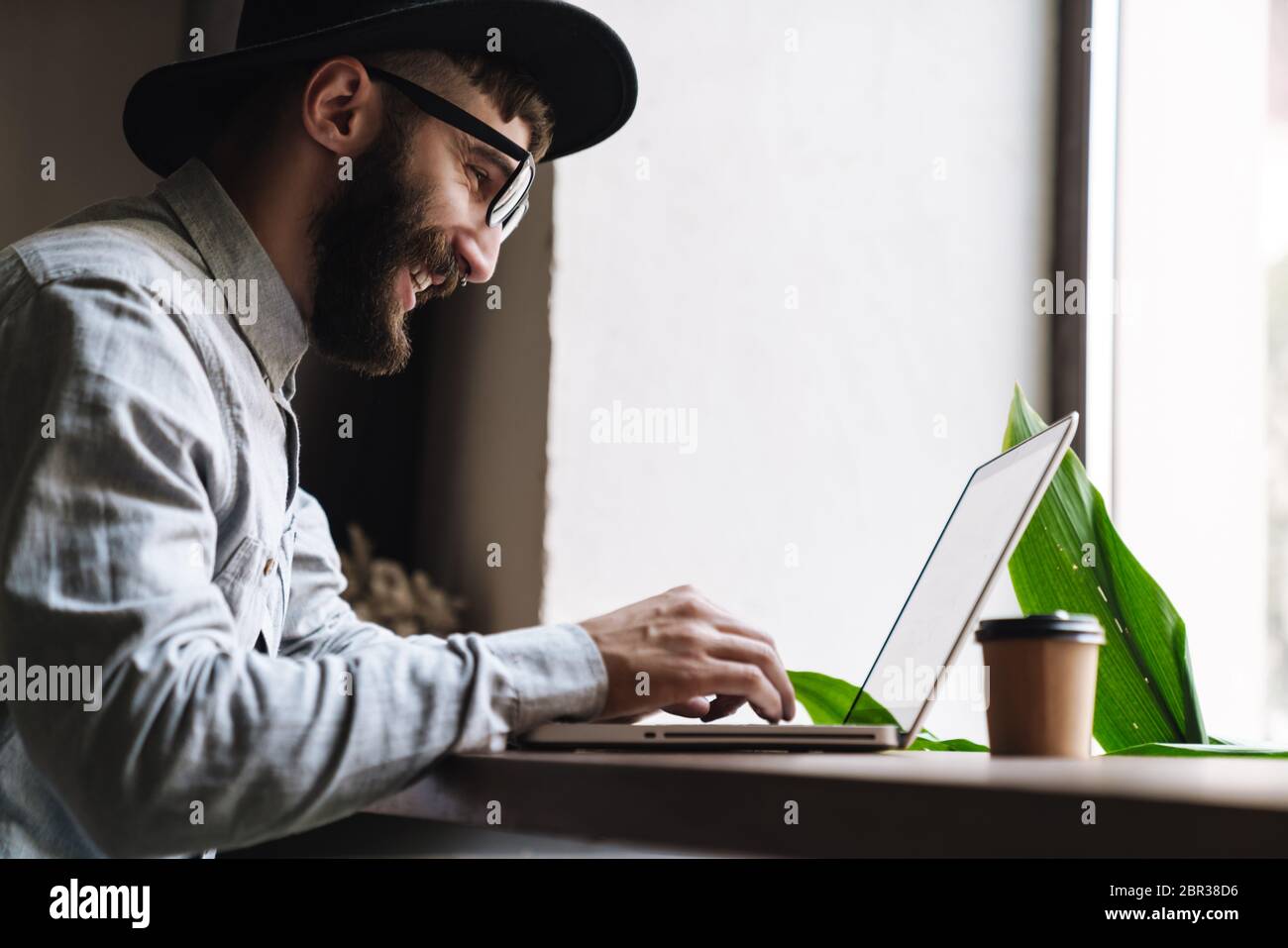 Photo of laughing young man wearing eyeglasses typing on laptop and ...