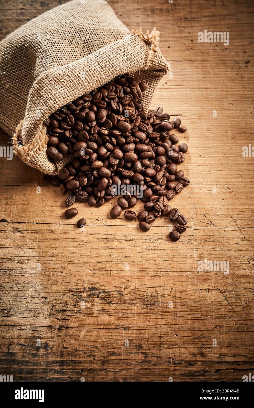 Full roasted blended coffee beans spilling from a small burlap bag onto rustic wood with copy ...