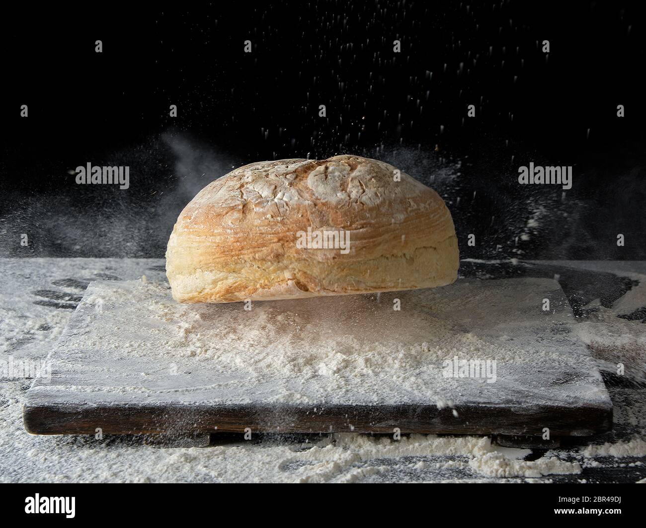 round baked bread in sprinkles of white wheat flour on a black ...