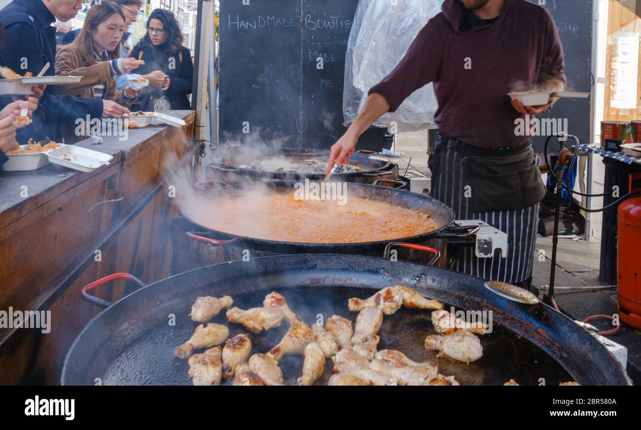 A man serves paella into a container next to large pans of cooking ...