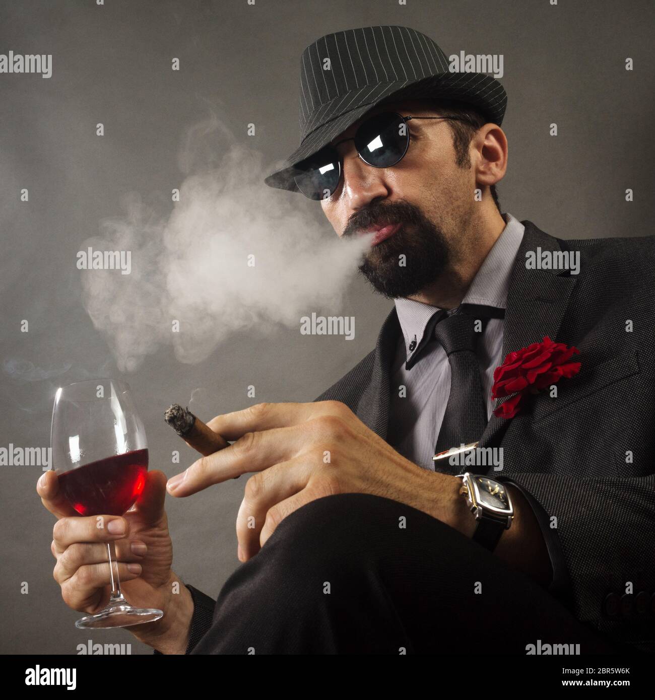 The elegant gentleman in suit with rose is smoking cigar and drinking ...