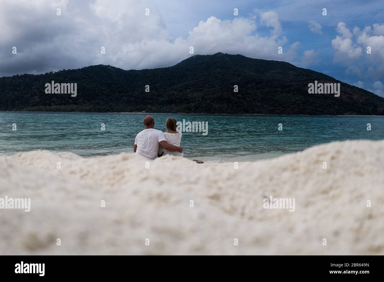 Back view couple sitting on the sandy beach and admiring the beautiful ...