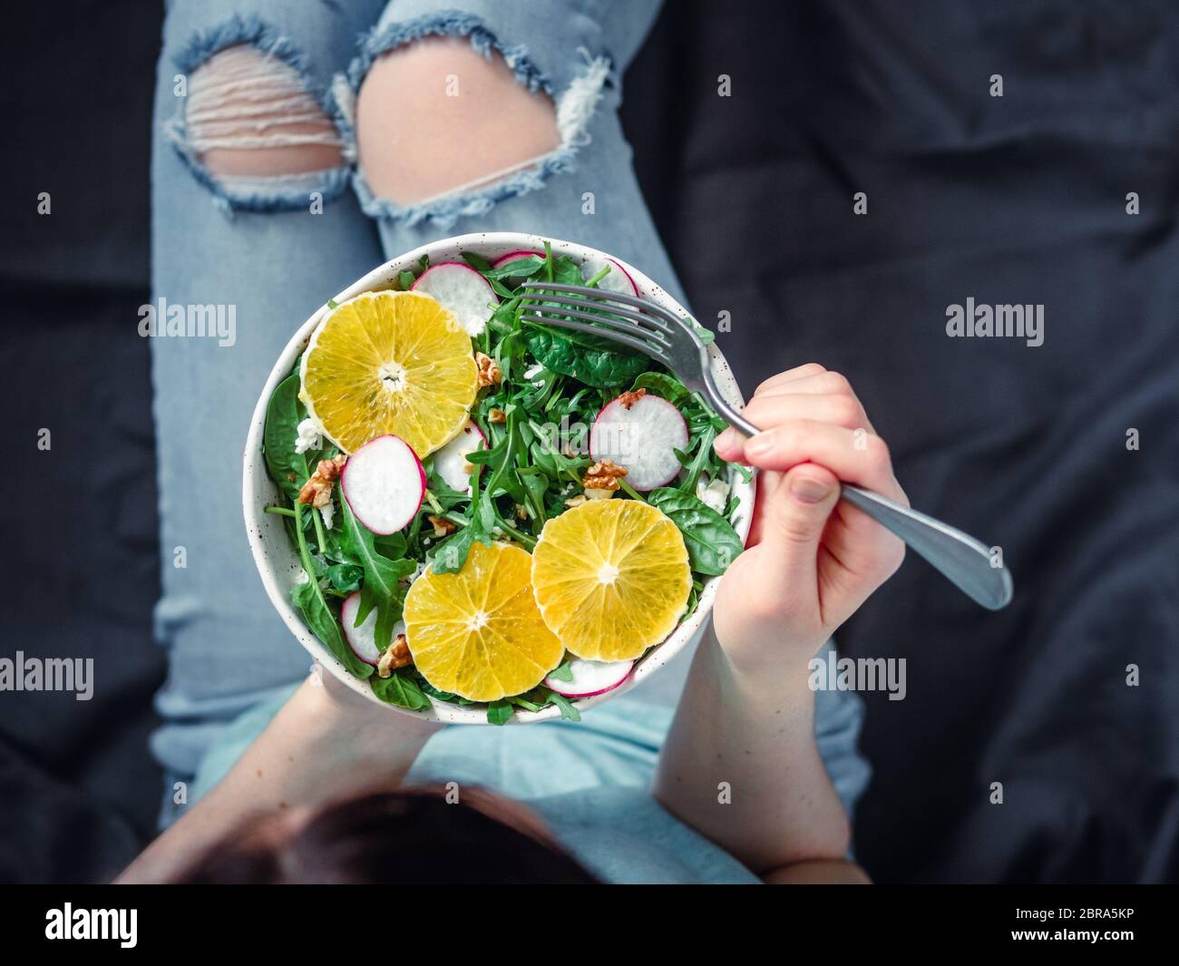 Woman in jeans at bed, holding vegan salad bowl with oranges, spinach ...