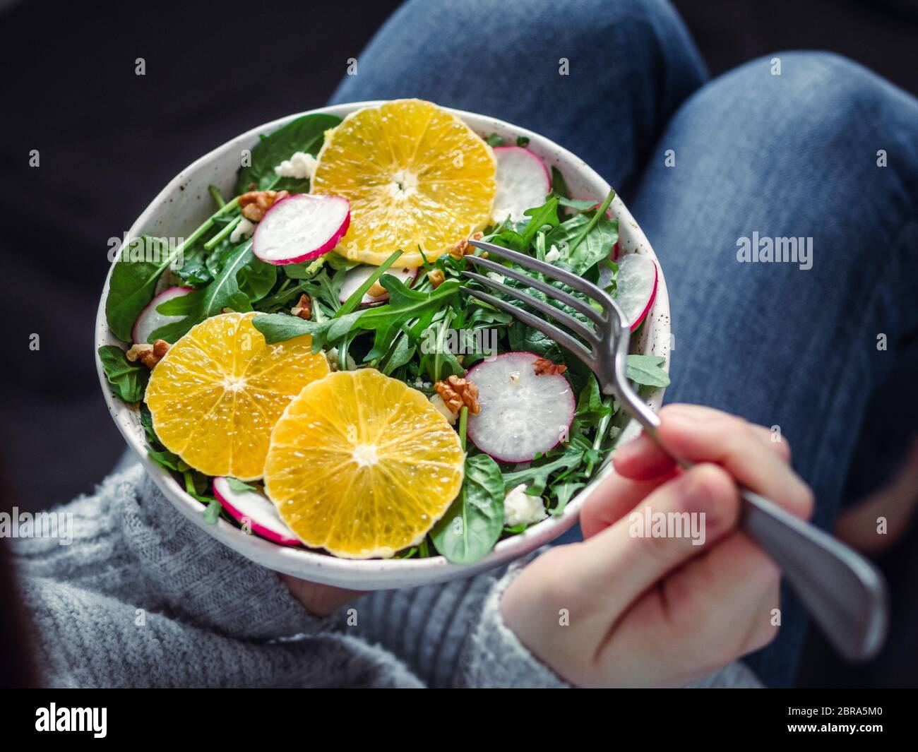 Woman in jeans at bed, holding vegan salad bowl with oranges, spinach ...