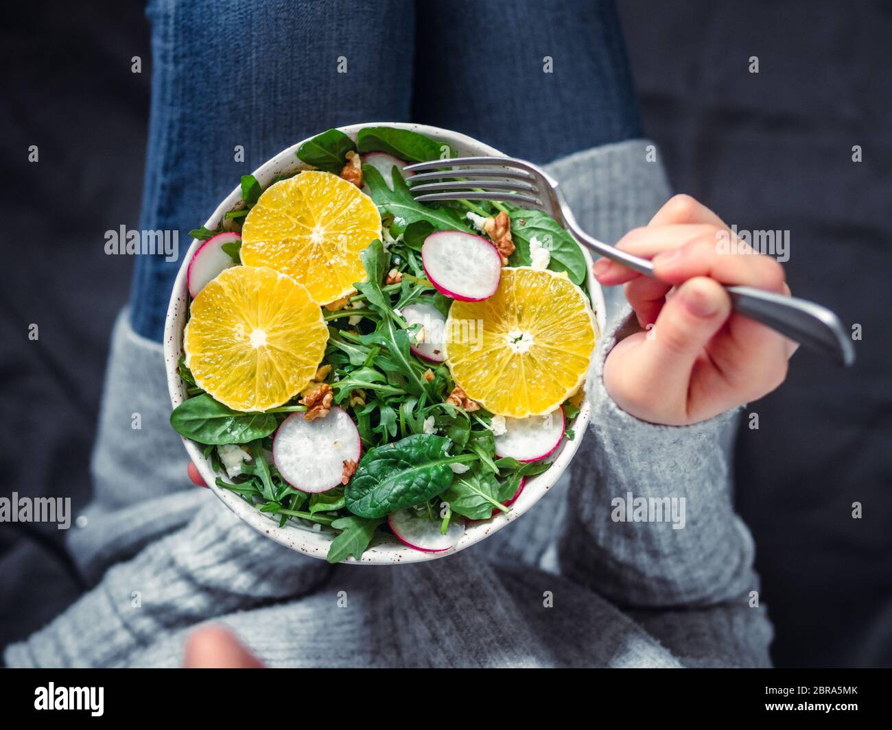Woman in jeans at bed, holding vegan salad bowl with oranges, spinach ...