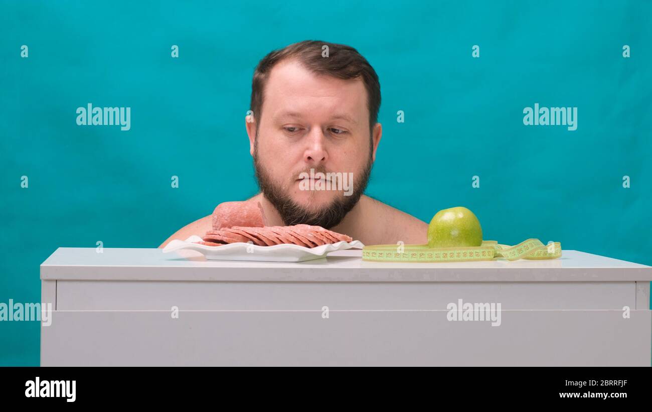 dull bearded fat man chooses between a sausage or a green apple on a ...