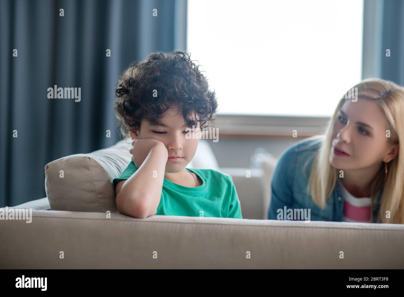 Sad curly boy sitting on sofa, blonde female talking to him, trying to ...
