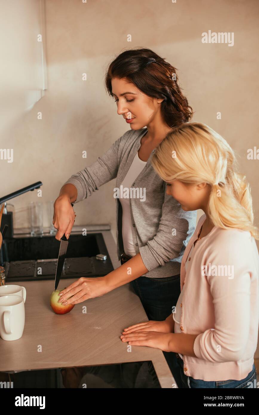 Side view of daughter looking at mother cutting apple on kitchen ...