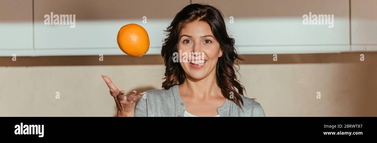 Panoramic crop of smiling woman throwing orange in kitchen Stock Photo ...