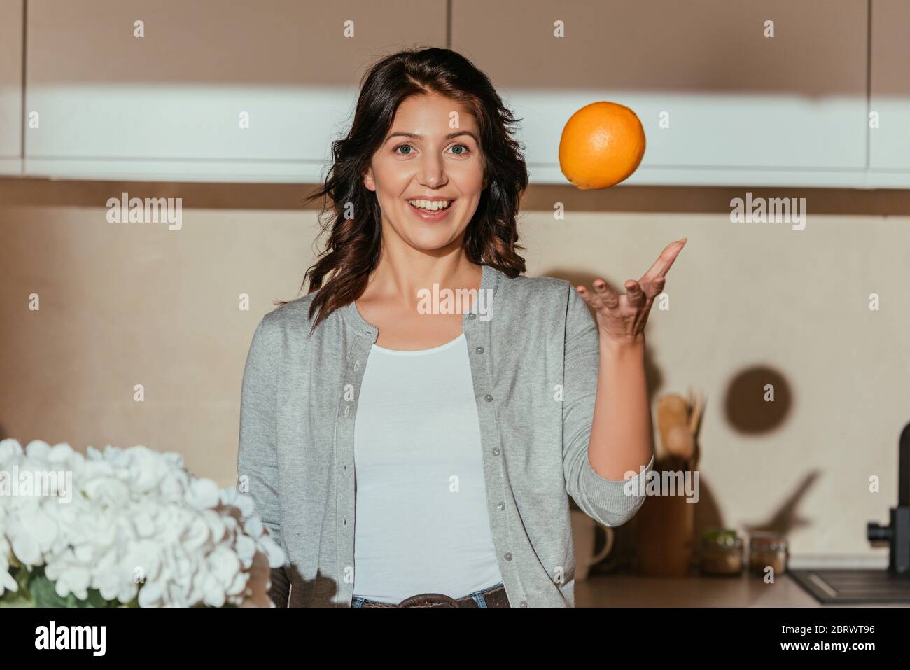 Selective focus of smiling woman throwing orange near flowers in ...