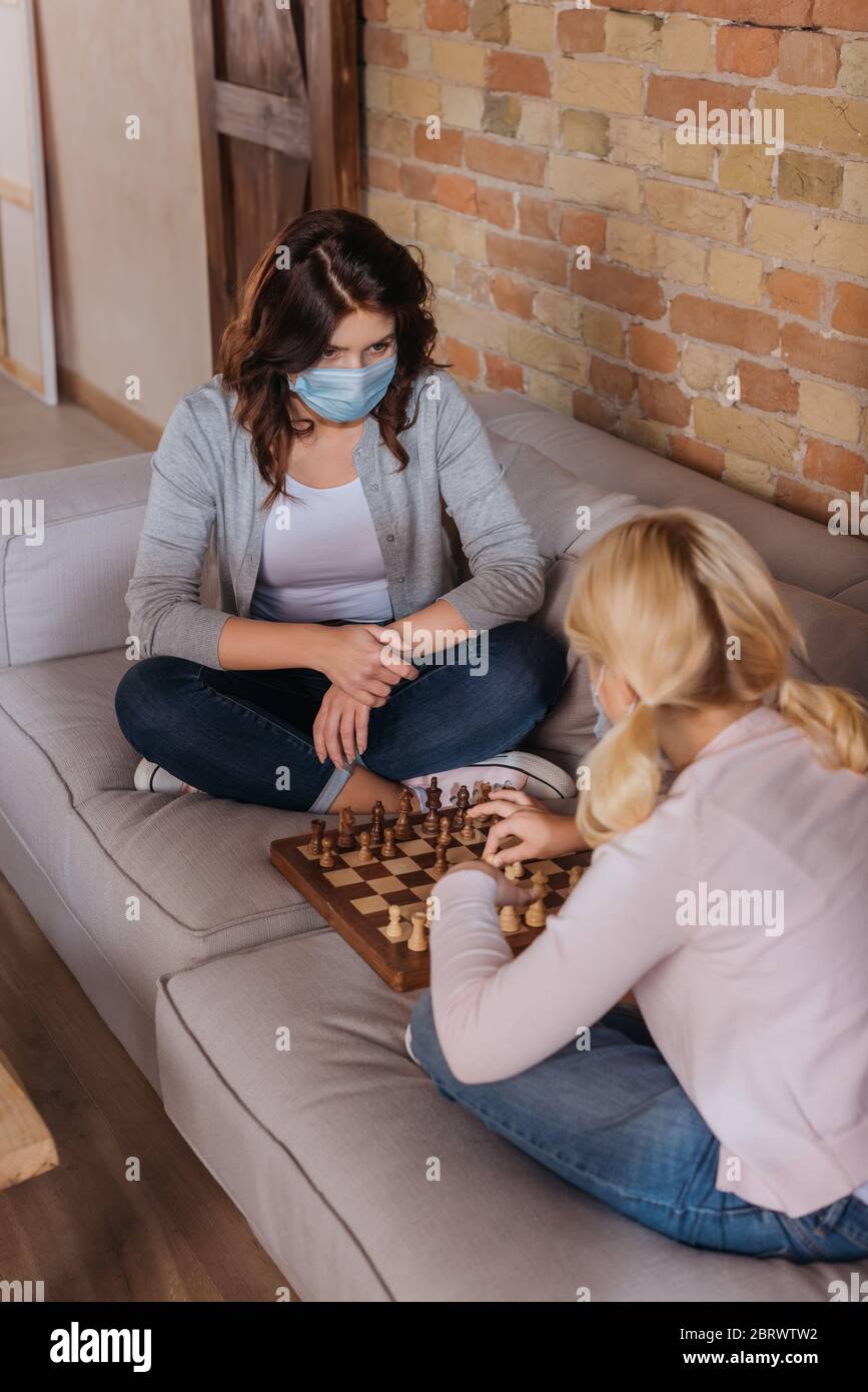 Woman in medical mask playing chess with child on couch in living room ...