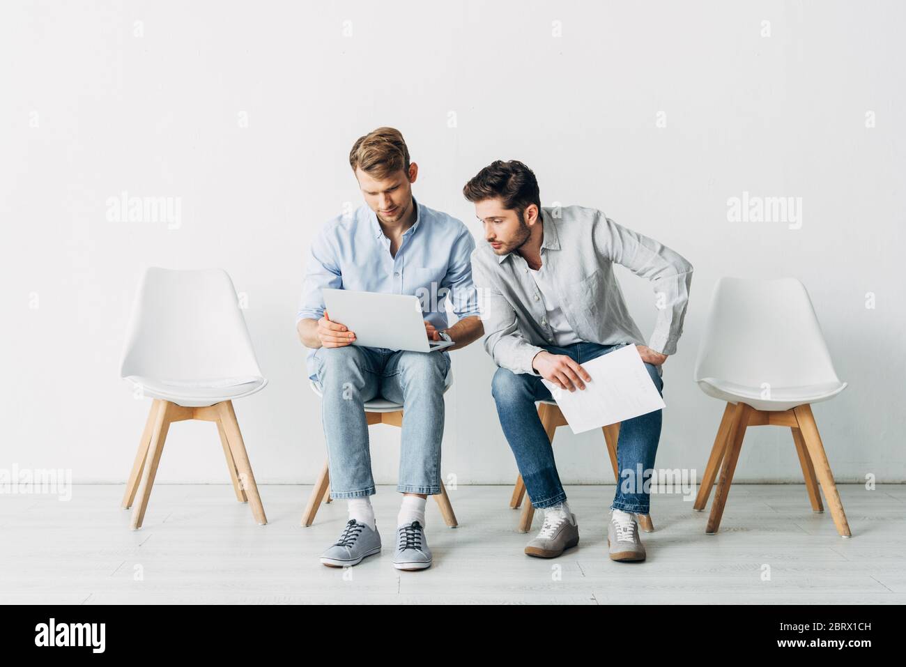 Two men with resume and laptop waiting for job interview on chairs in ...
