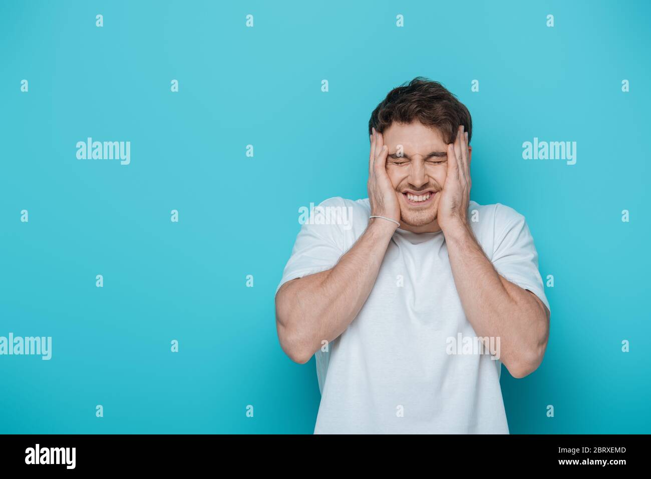 scared young man touching face while grimacing with closed eyes on blue ...