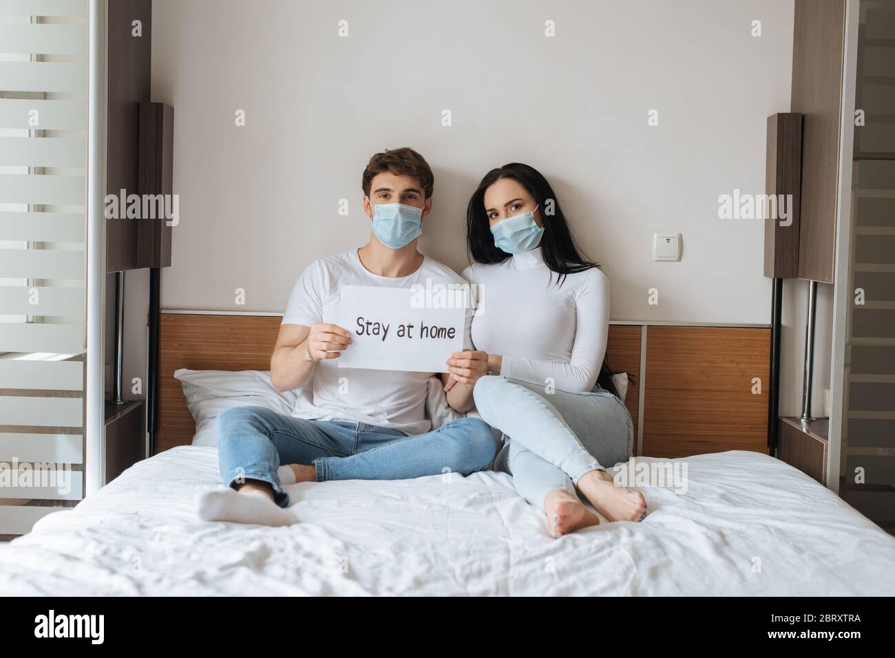 young couple in medical masks holding Stay at home sign in bedroom ...
