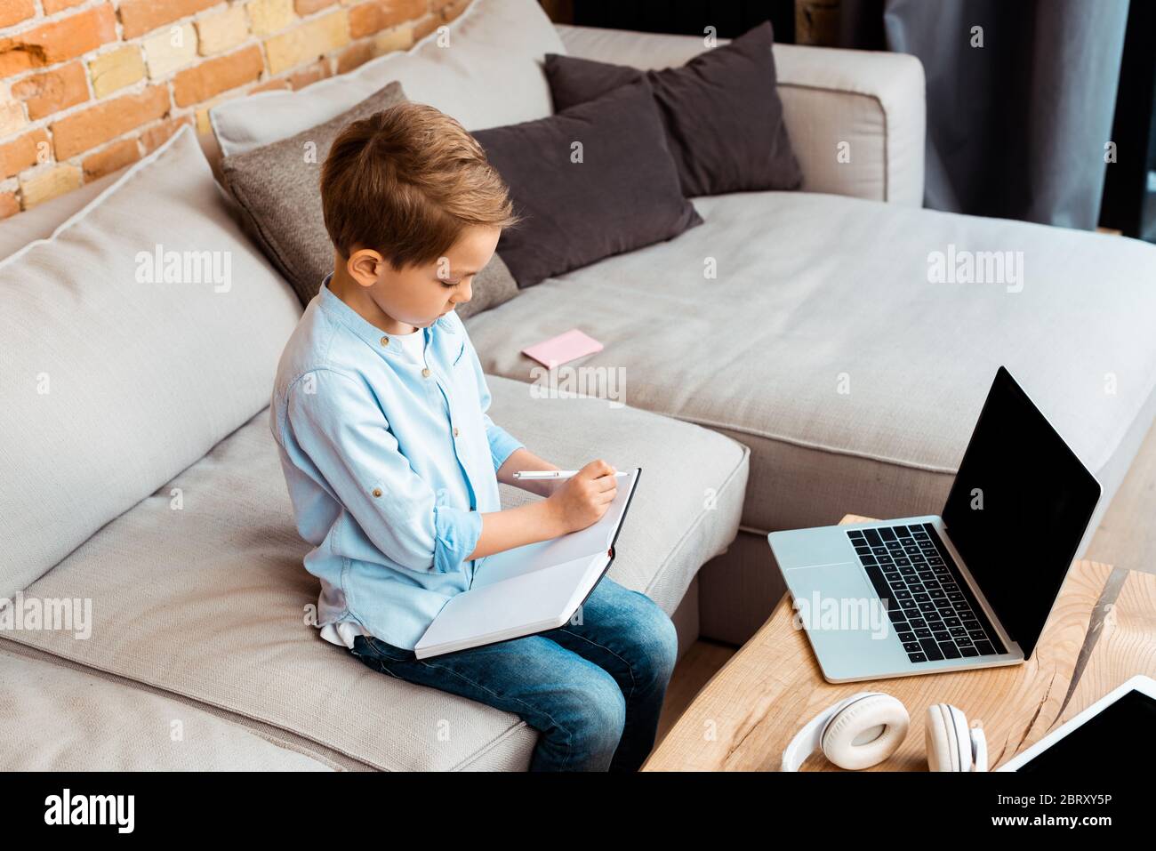 Adorable Boy Writing In Notebook Near Gadgets With Blank Screen While E adorable-boy-writing-in-notebook-near-gadgets-with-blank-screen-while-e