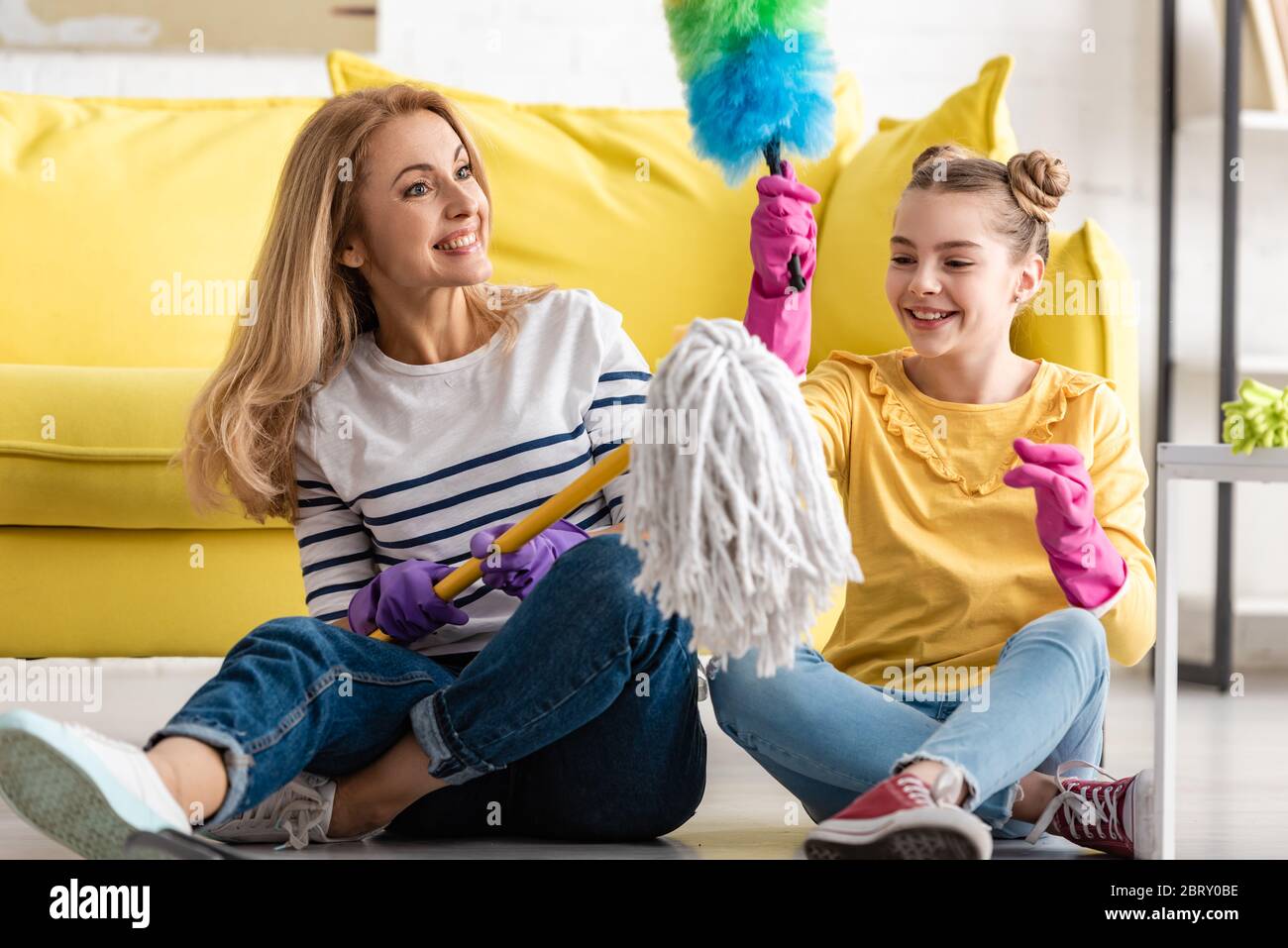 Mother and cute daughter with mop and feather duster smiling on floor ...