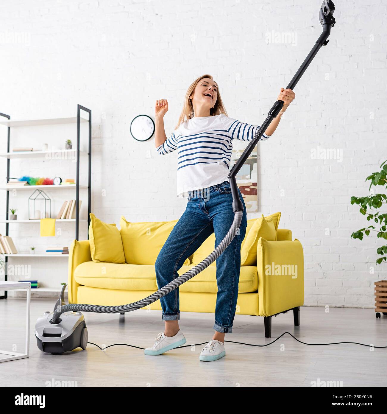 Blonde woman singing with closed eyes and vacuum cleaner near sofa in ...