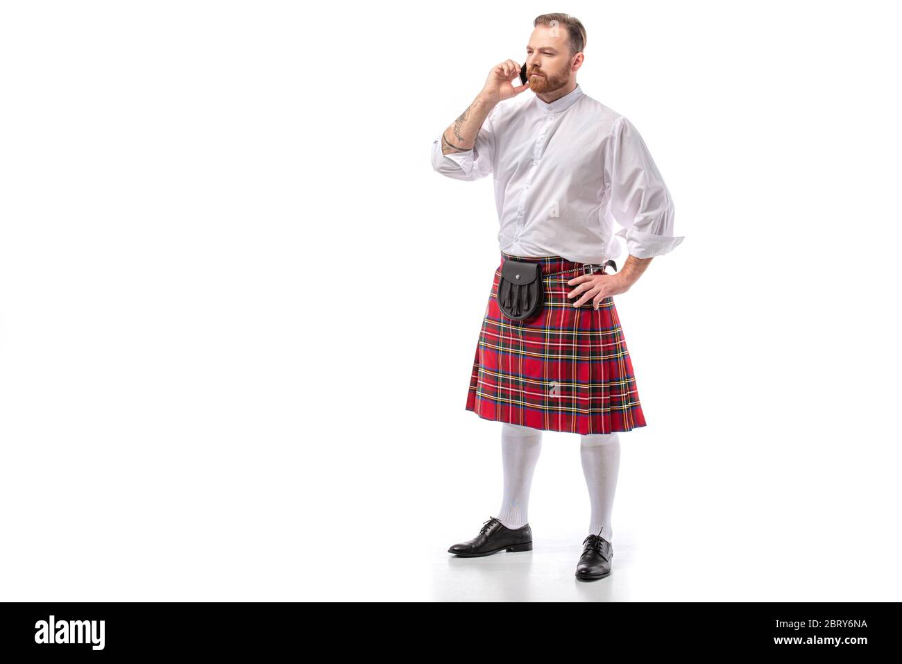 serious Scottish redhead man in red kilt talking on smartphone on white ...