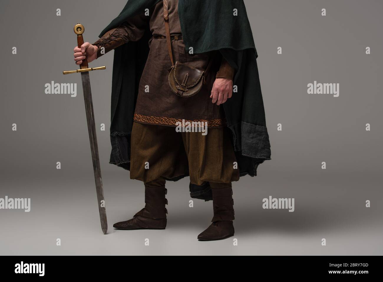 cropped view of medieval Scottish knight in mantel with sword on grey ...