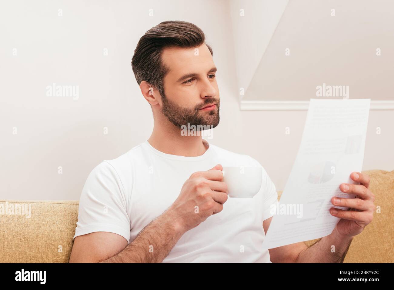 Handsome teleworker holding document and drinking coffee on couch Stock ...