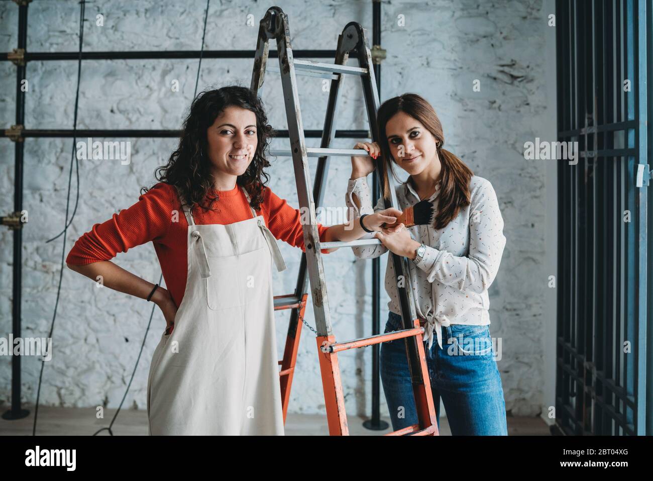 Two mid adult women leaning on step ladders with paint brush in their ...