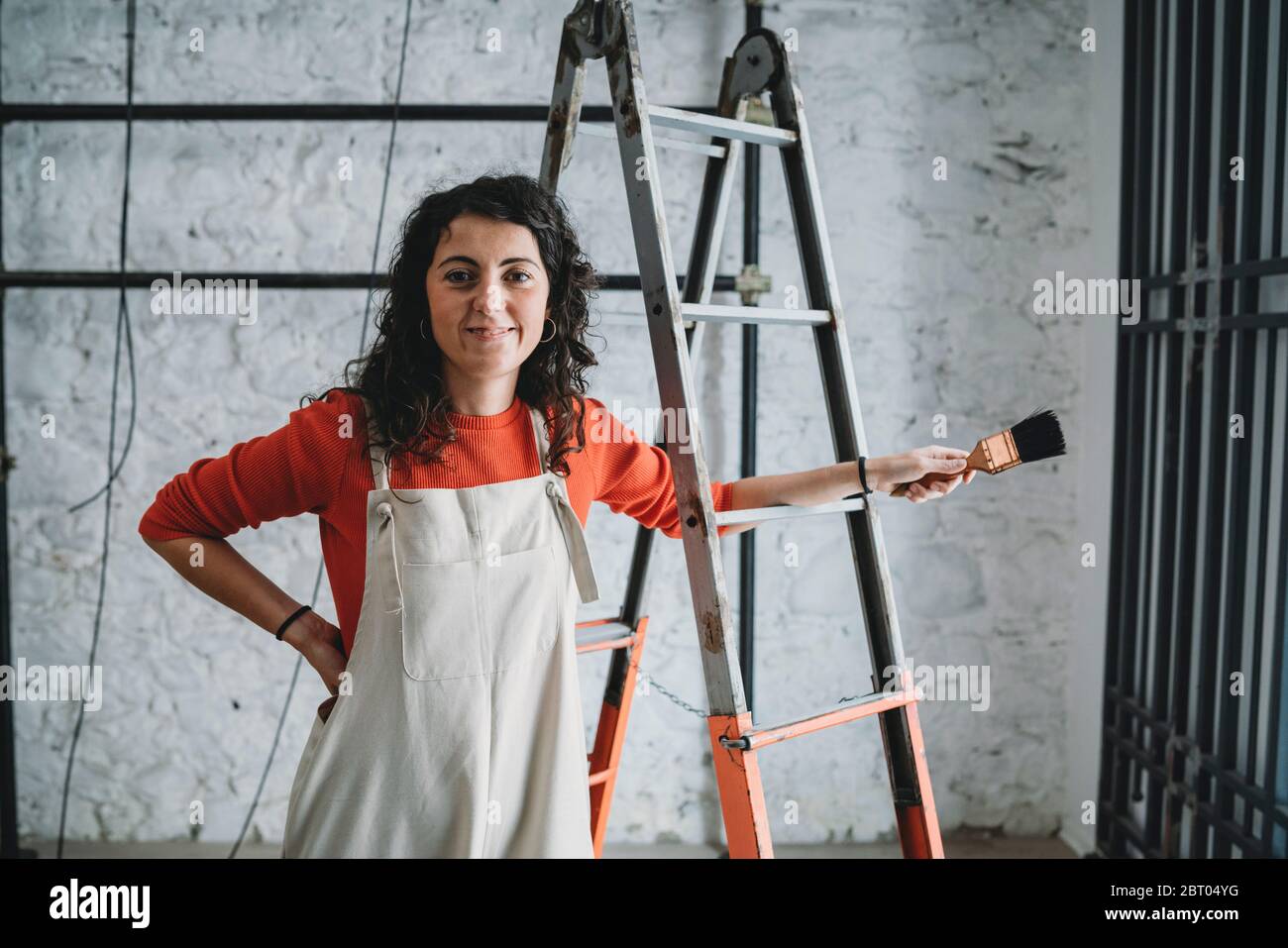 Mid adult woman leaning on step ladder with paint brush in her new shop ...