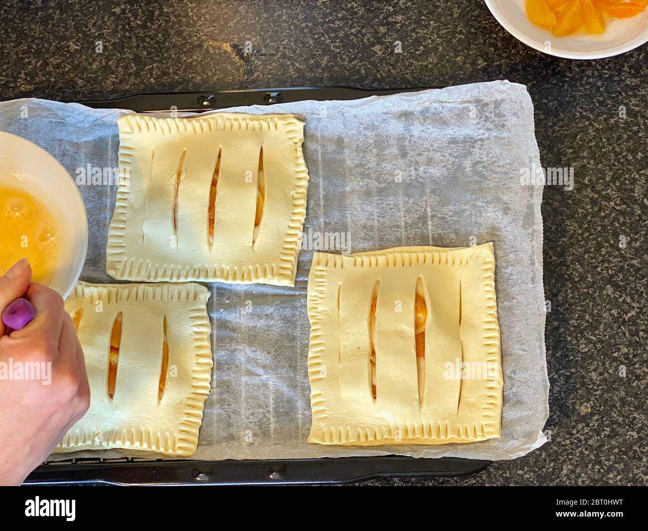 Hand applying egg on uncooked apricot puff pastry on a baking tray in a ...
