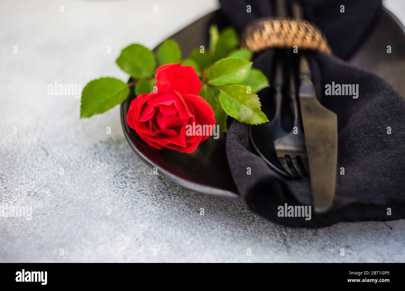 Table setting with fresh red rose flower and black plate and cutlery on ...