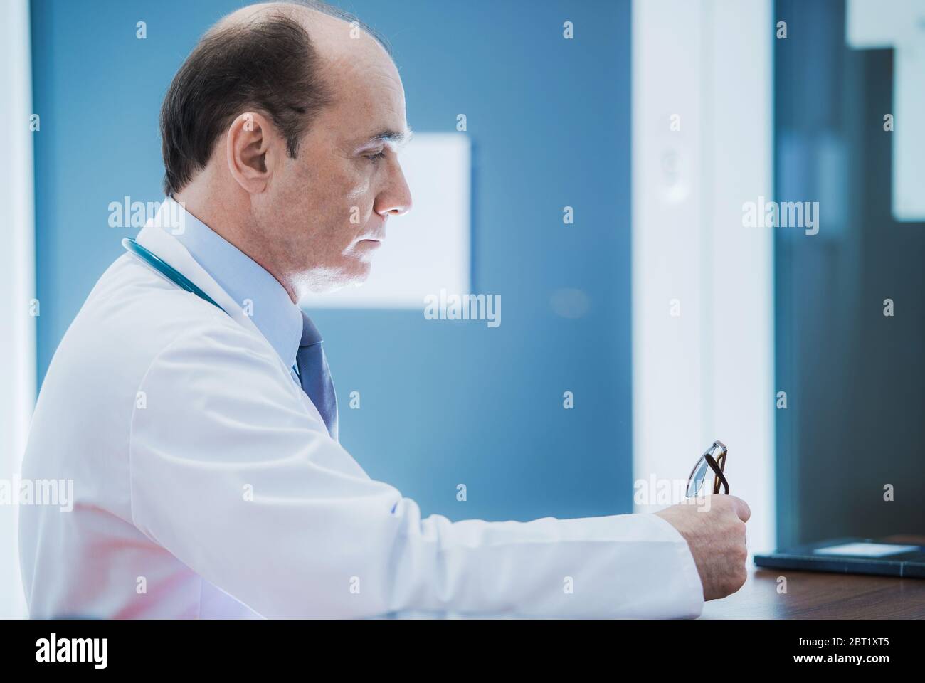 Caucasian Male Doctor In Lab Coat Holding Glasses Sitting At Desk In ...