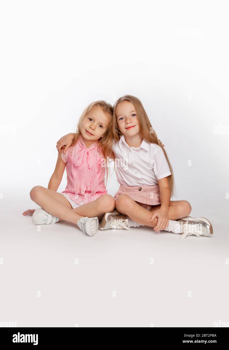 Two cute girlfriends are sitting hugging and legs crossed on the floor ...