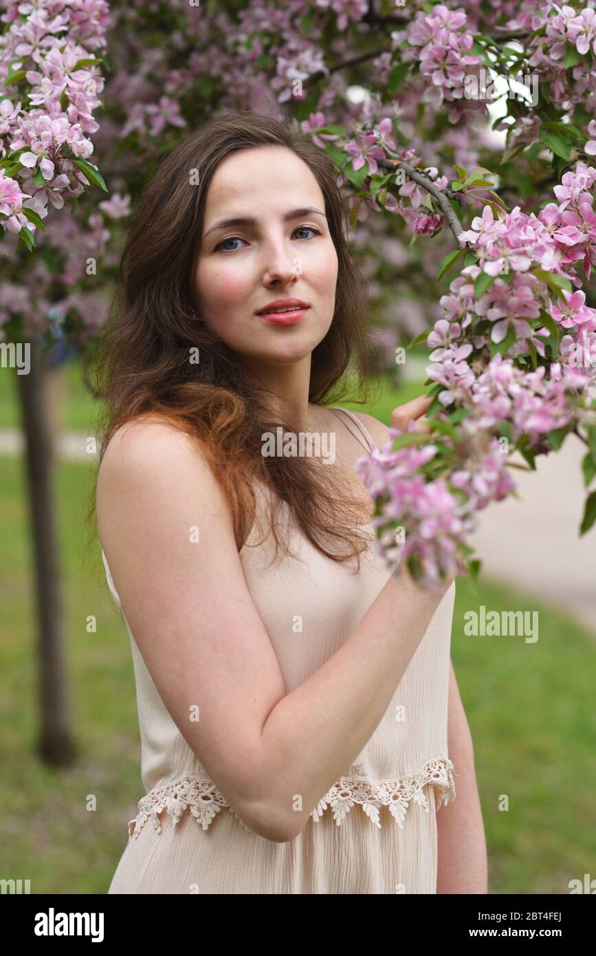 A young beautiful woman with a slight smile touches a blooming branch ...