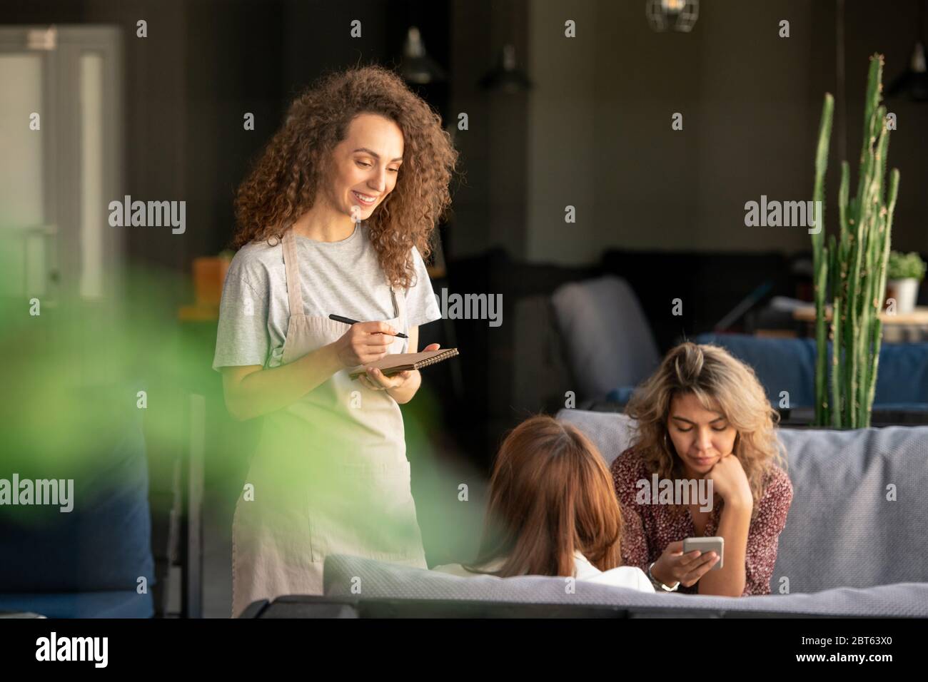 Happy young brunette waitress in apron looking at one of clients and ...
