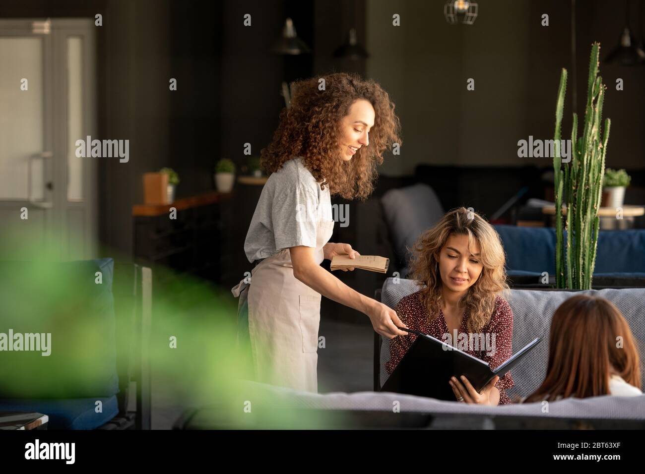 Young friendly waitress of restaurant pointing at menu while giving ...